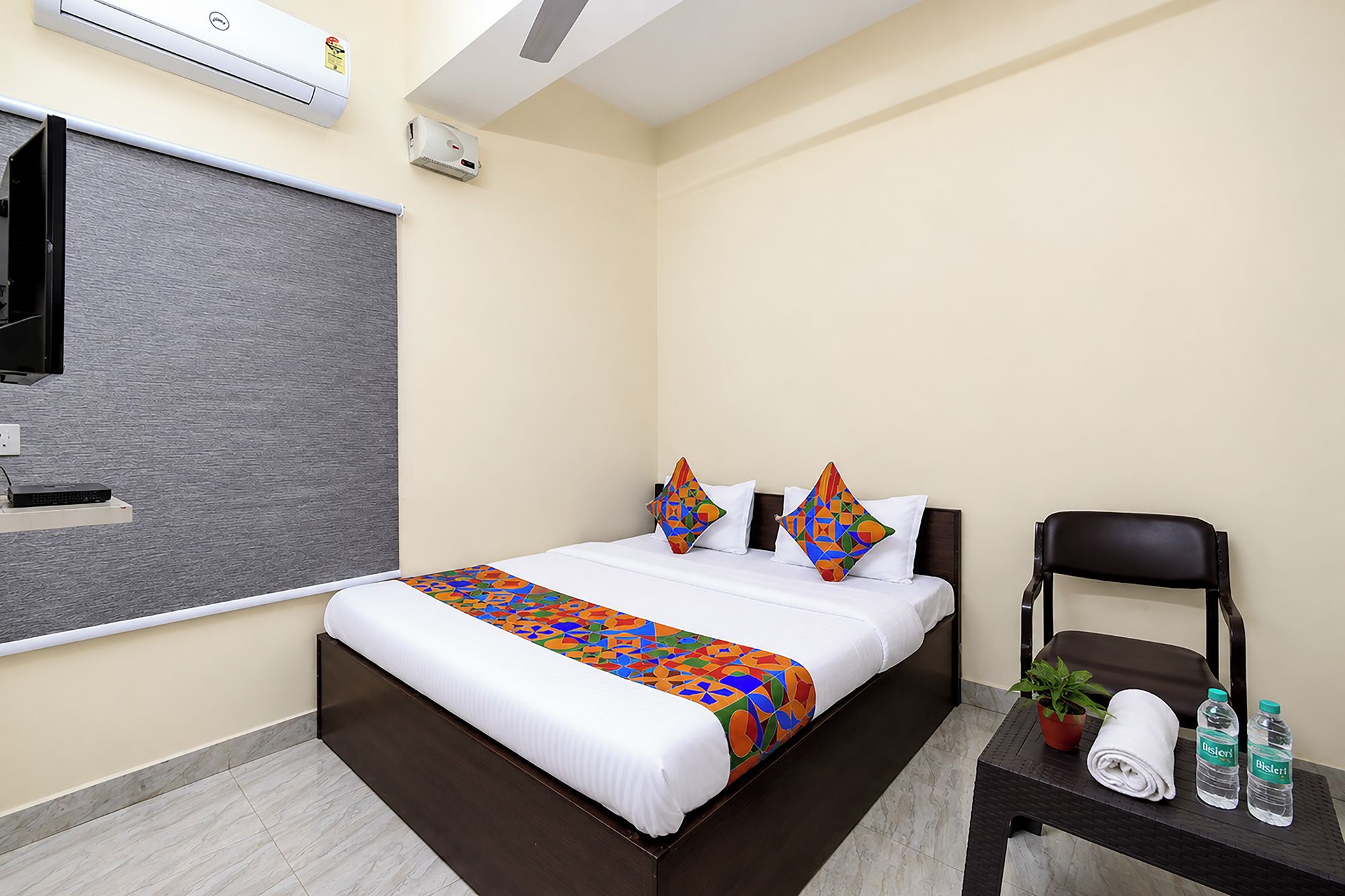 Quarto de hotel com uma cama, uma escrivaninha, uma cadeira, um vaso de plantas e uma garrafa de água.