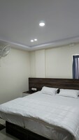 Quarto com cama, mesa de cabeceira, ventoinha na parede e iluminação embutida no teto.