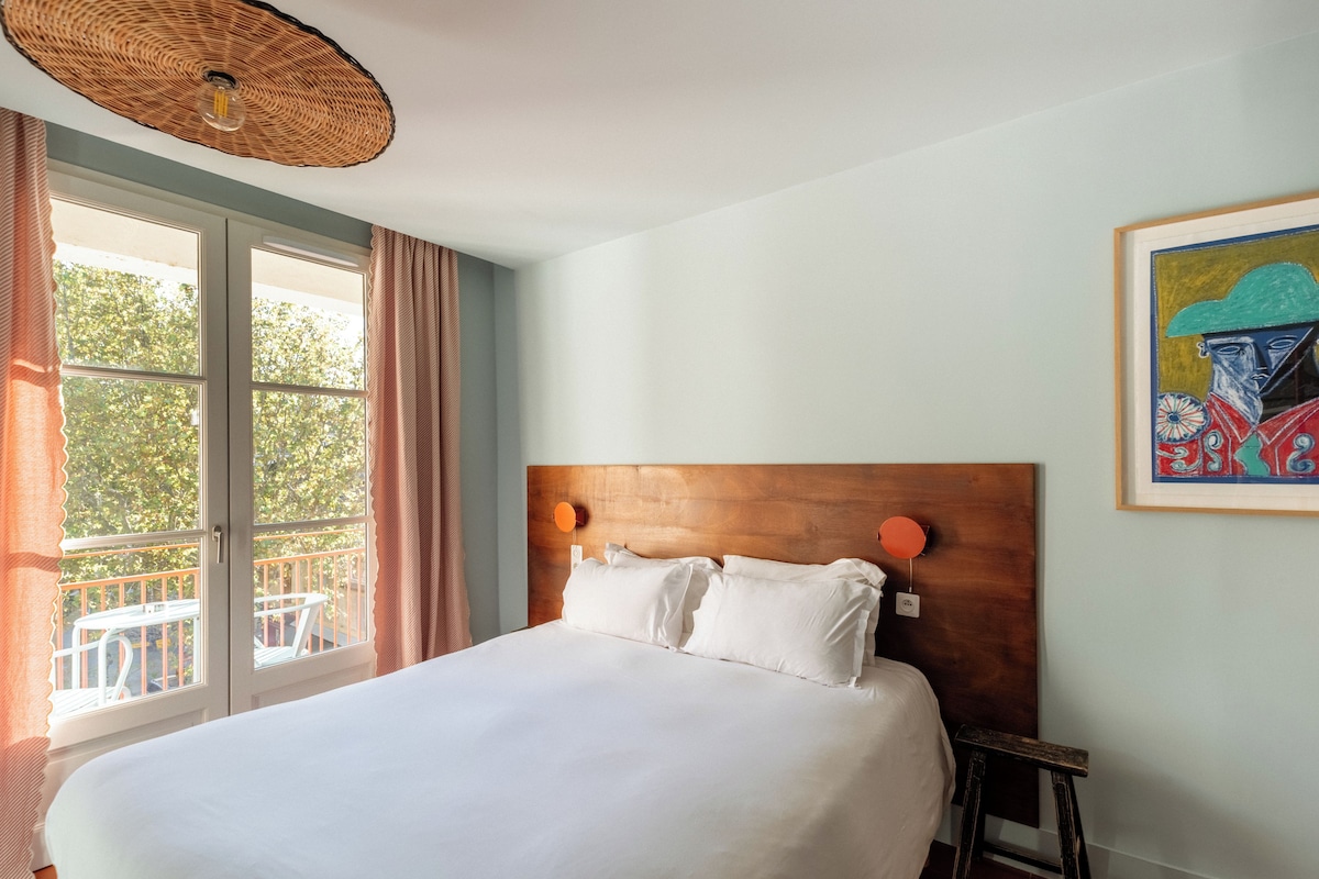 Quarto de hotel com uma cama grande, cabeceira de madeira, um quadro na parede e varanda com vista para árvores.