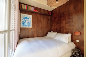 Quarto com cama grande, um banquinho, estante com livros e quadro na parede.