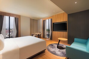 Quarto de hotel com cama, área de estar com mesa e cadeiras, TV de tela plana na parede e janelas amplas com vista para a cidade.
