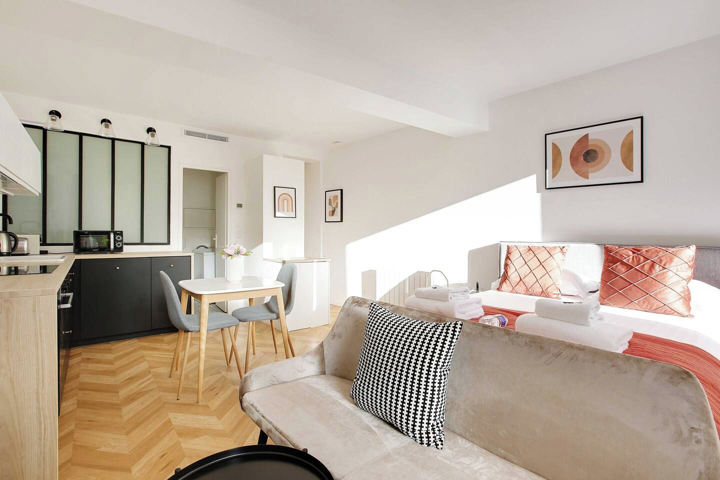 Quarto de hotel moderno com sofá, cama com almofadas decorativas, área de jantar com mesa e cadeiras, e cozinha compacta com micro-ondas.