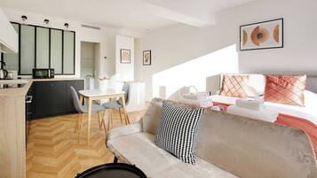Quarto de hotel moderno com sofá, cama com almofadas decorativas, área de jantar com mesa e cadeiras, e cozinha compacta com micro-ondas.
