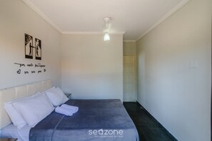 Quarto com cama, almofadas brancas, manta cinza, ventilador de teto e parede com objeto decorativo e texto.