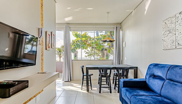Uma sala de estar moderna com TV de tela plana, um sofá azul, uma mesa de jantar com cadeiras e uma grande janela com cortinas.