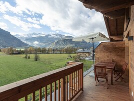 Um deck de madeira com mesa e cadeiras, com vista para um campo verde e montanhas.