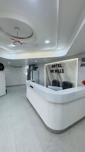 Uma área de recepção de hotel com balcão de mármore, ventoinha no teto e uma placa com a inscrição 'HOTEL HR HILLS'.