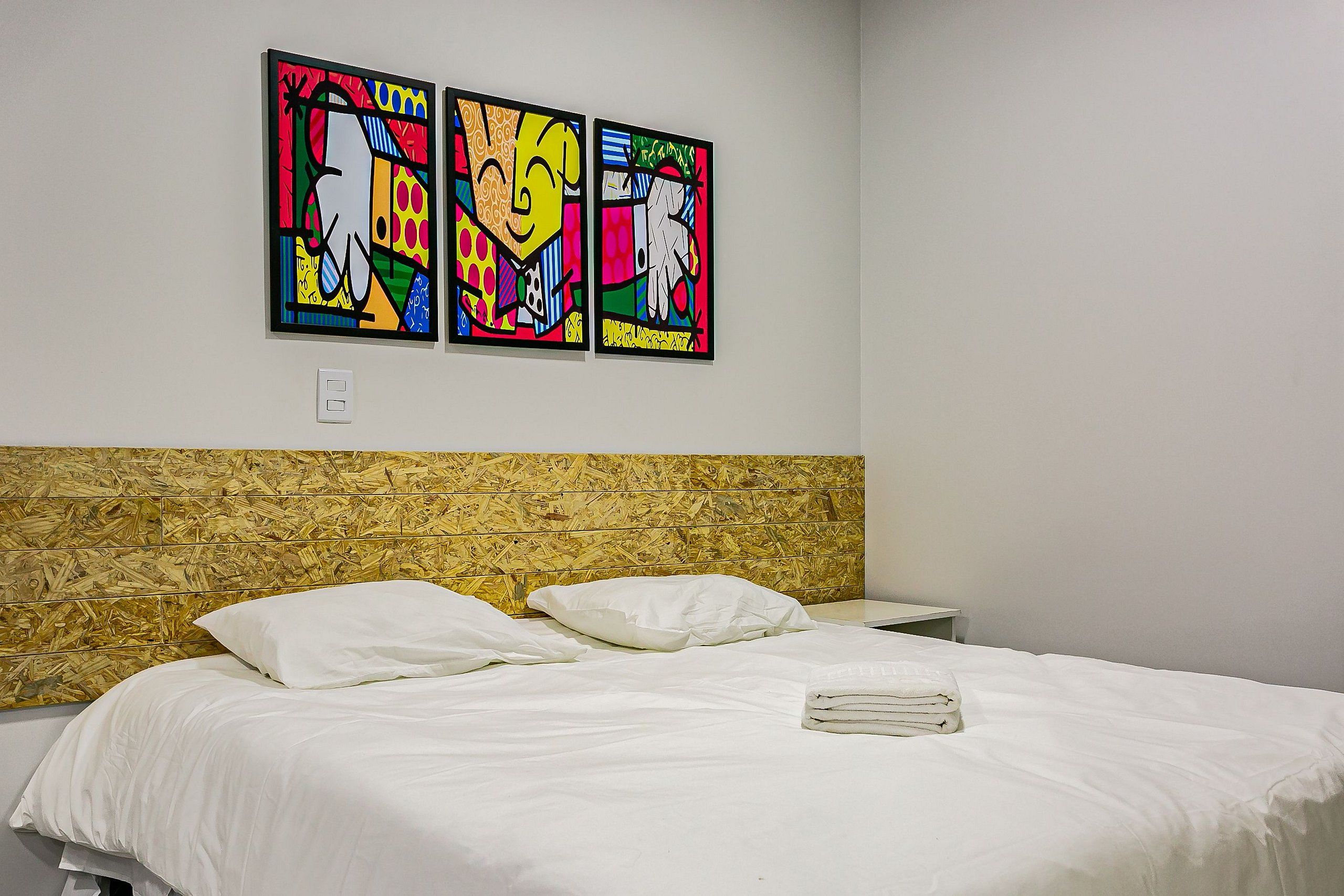 Quarto de hotel com cabeceira de madeira, cama com roupa de cama branca e duas pinturas abstratas coloridas na parede.