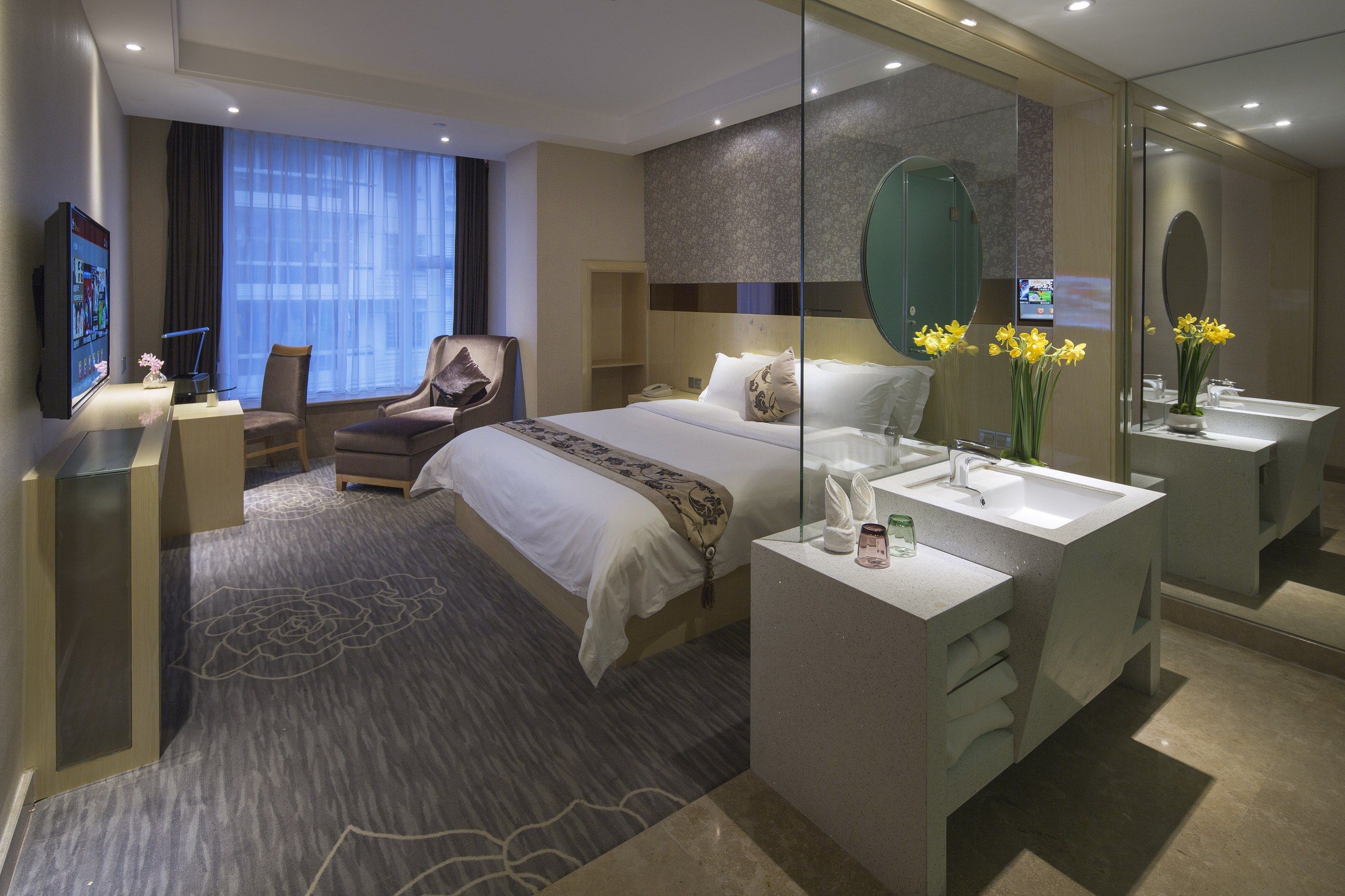Quarto de hotel com cama, escrivaninha com televisão de tela plana, cadeira, espelho grande e banheiro com pia e espelho.