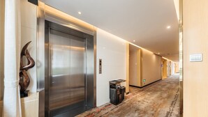 Corredor de hotel com um elevador parado, uma lixeira e piso coberto por carpete.