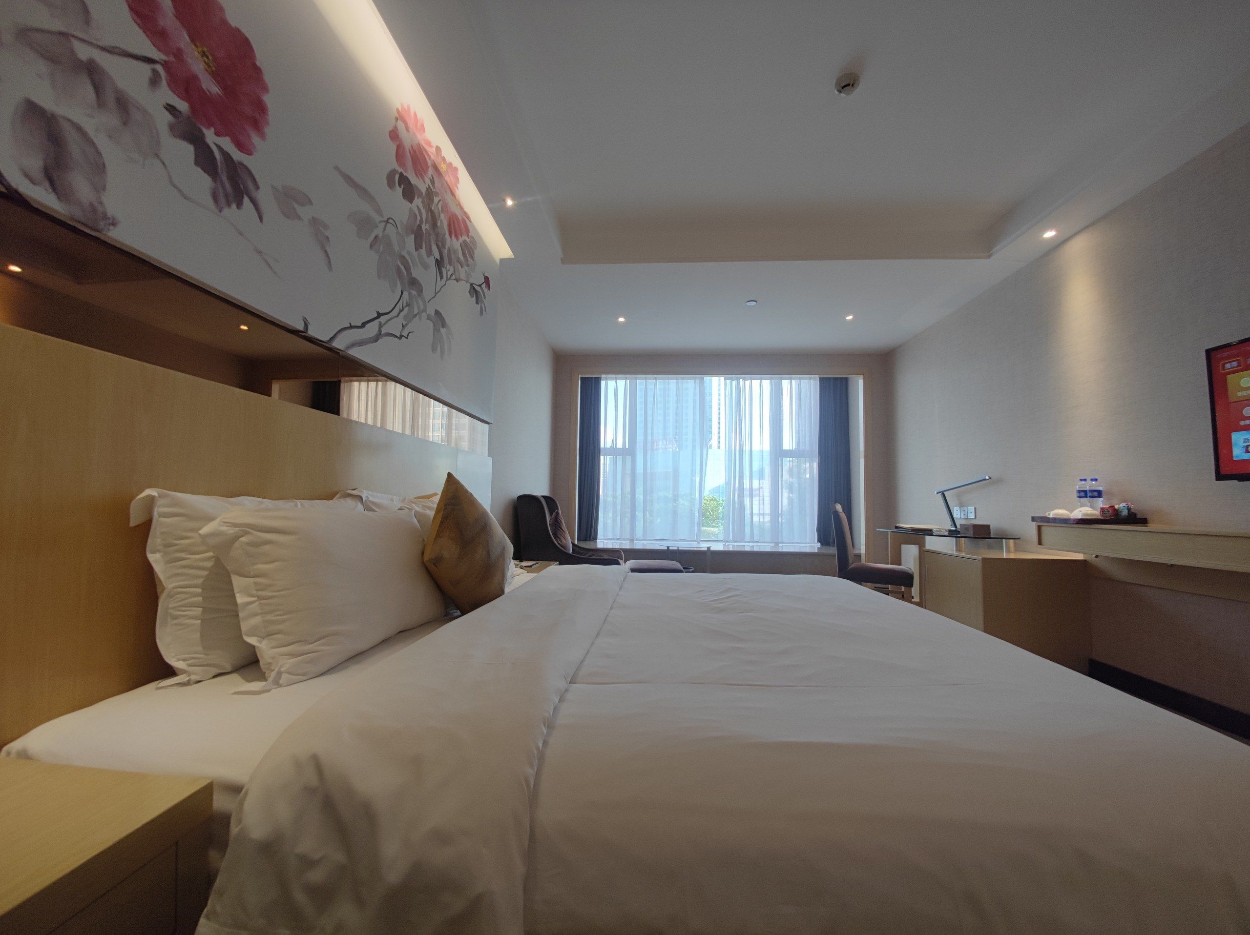 Quarto de hotel com uma cama grande, escrivaninha, cadeira e janela com cortinas.