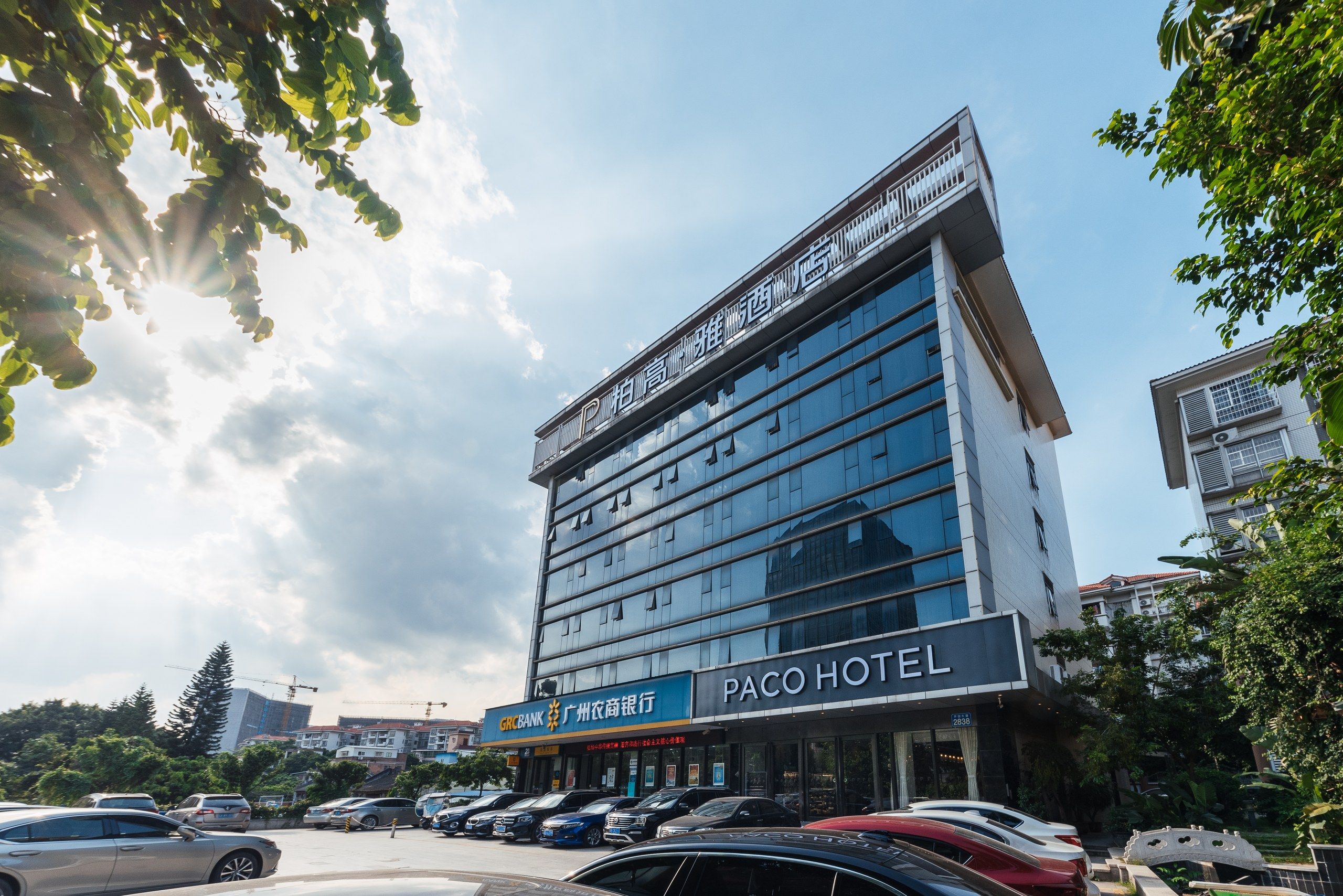 Um edifício de vários andares com fachada de vidro moderna, um restaurante visível no térreo chamado 'Paco Hotel' e um estacionamento com vários carros.