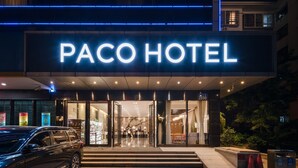 Entrada de um hotel com uma placa chamativa, um carro estacionado na frente e uma escadaria que leva à entrada.