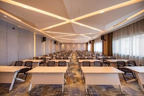 Sala de conferência com fileiras de mesas e cadeiras, carpete estampado e teto com iluminação linear.