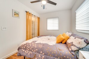 Quarto com cama, ventilador de teto, janela com persianas e interruptor de luz na parede.
