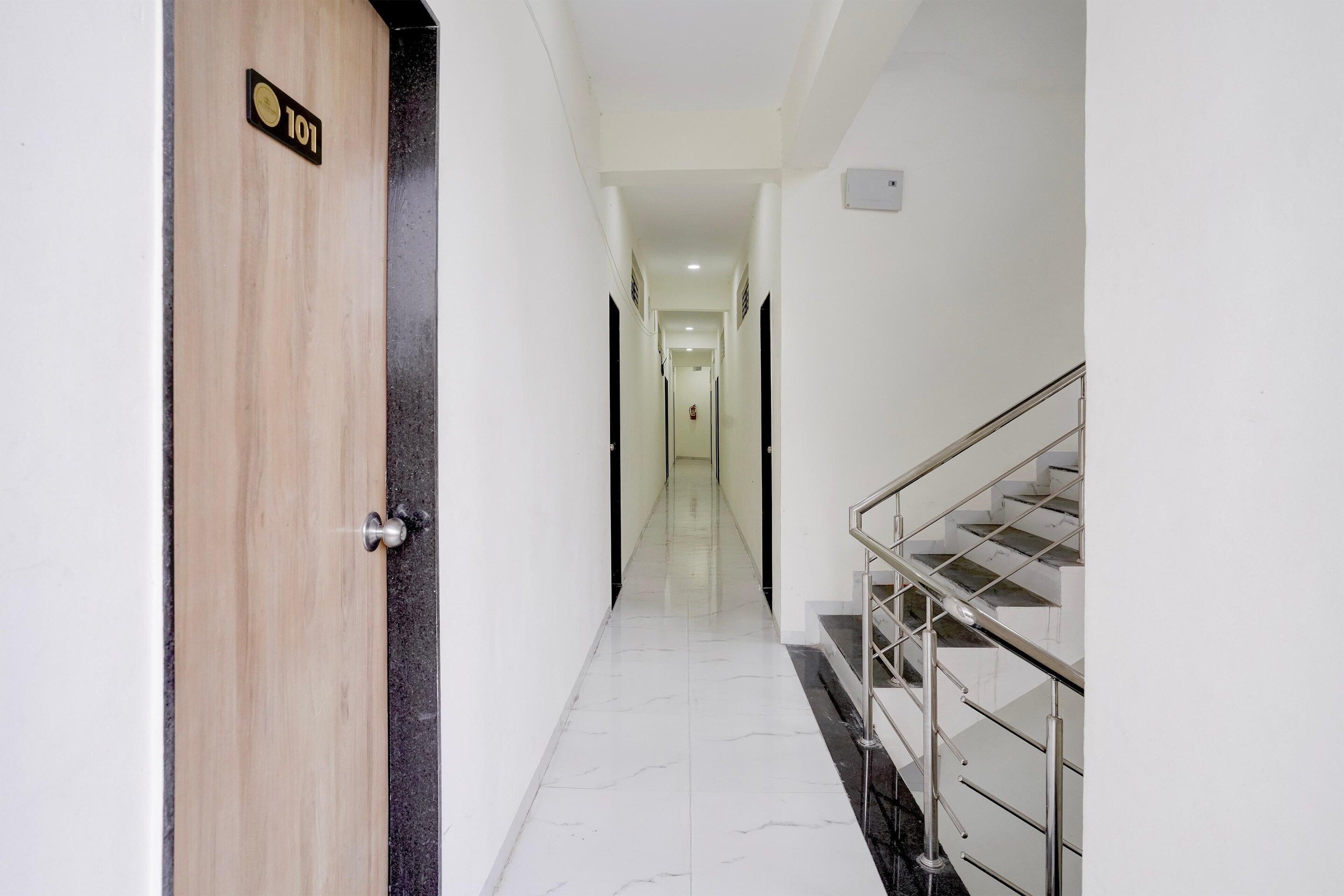 Corredor de hotel com porta de madeira identificada com o número '101', piso de mármore e escada com guarda-corpos de metal.