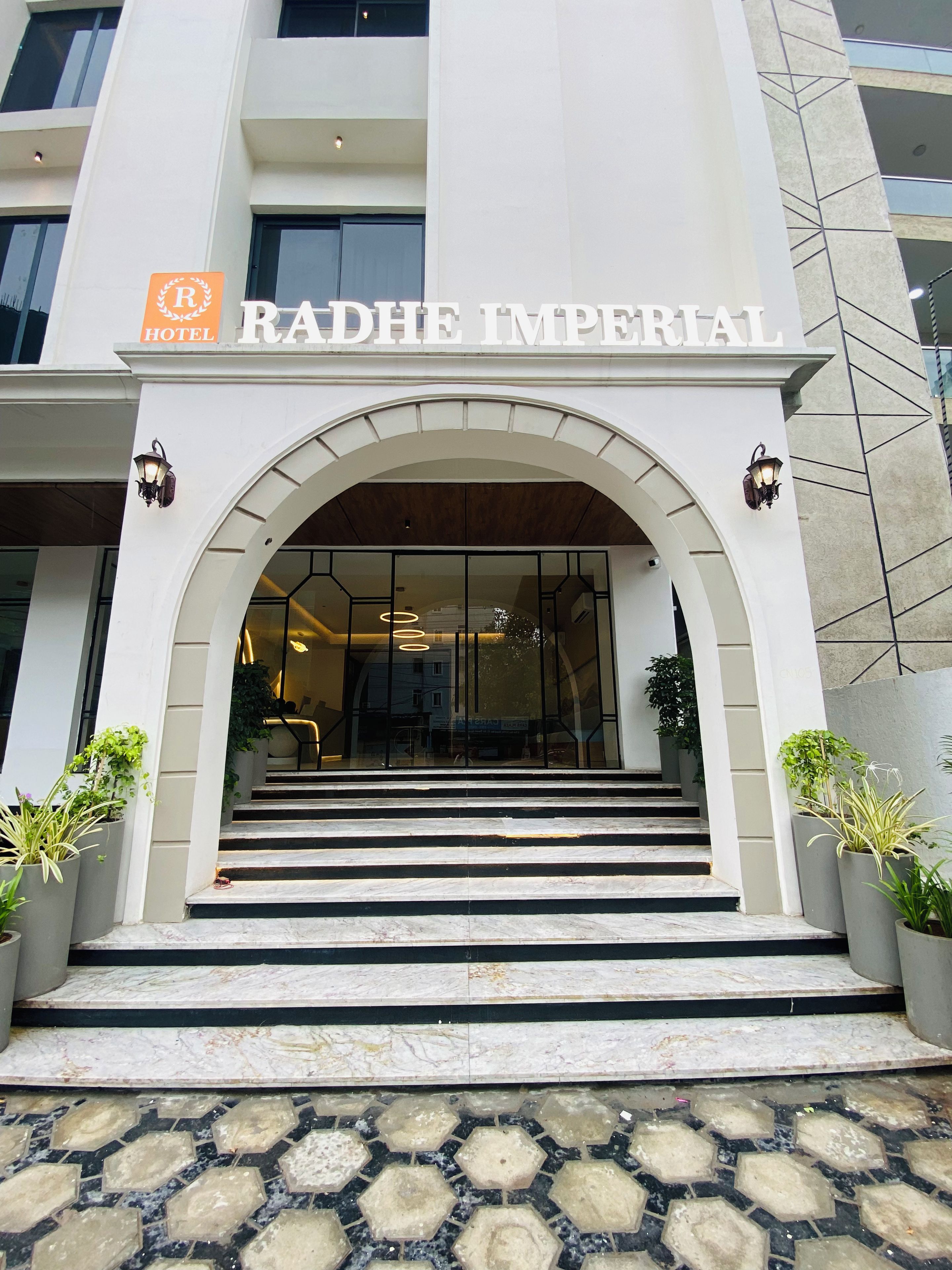 A entrada do Radhe Imperial Hotel, com um design moderno, apresenta uma grande porta em arco, degraus que levam à entrada e vasos de plantas de ambos os lados.