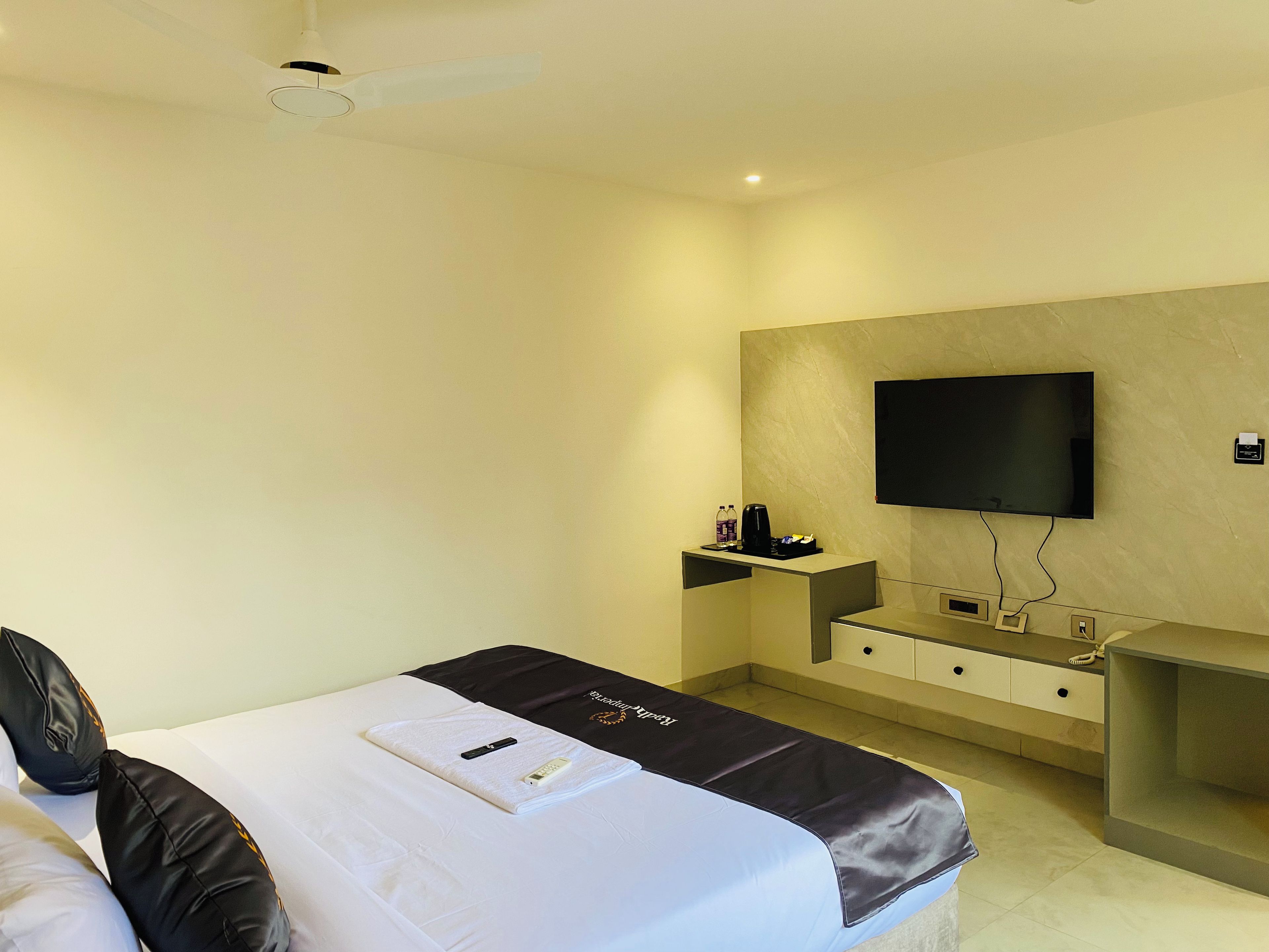 Quarto de hotel com cama, televisão na parede, escrivaninha e ventilador de teto.