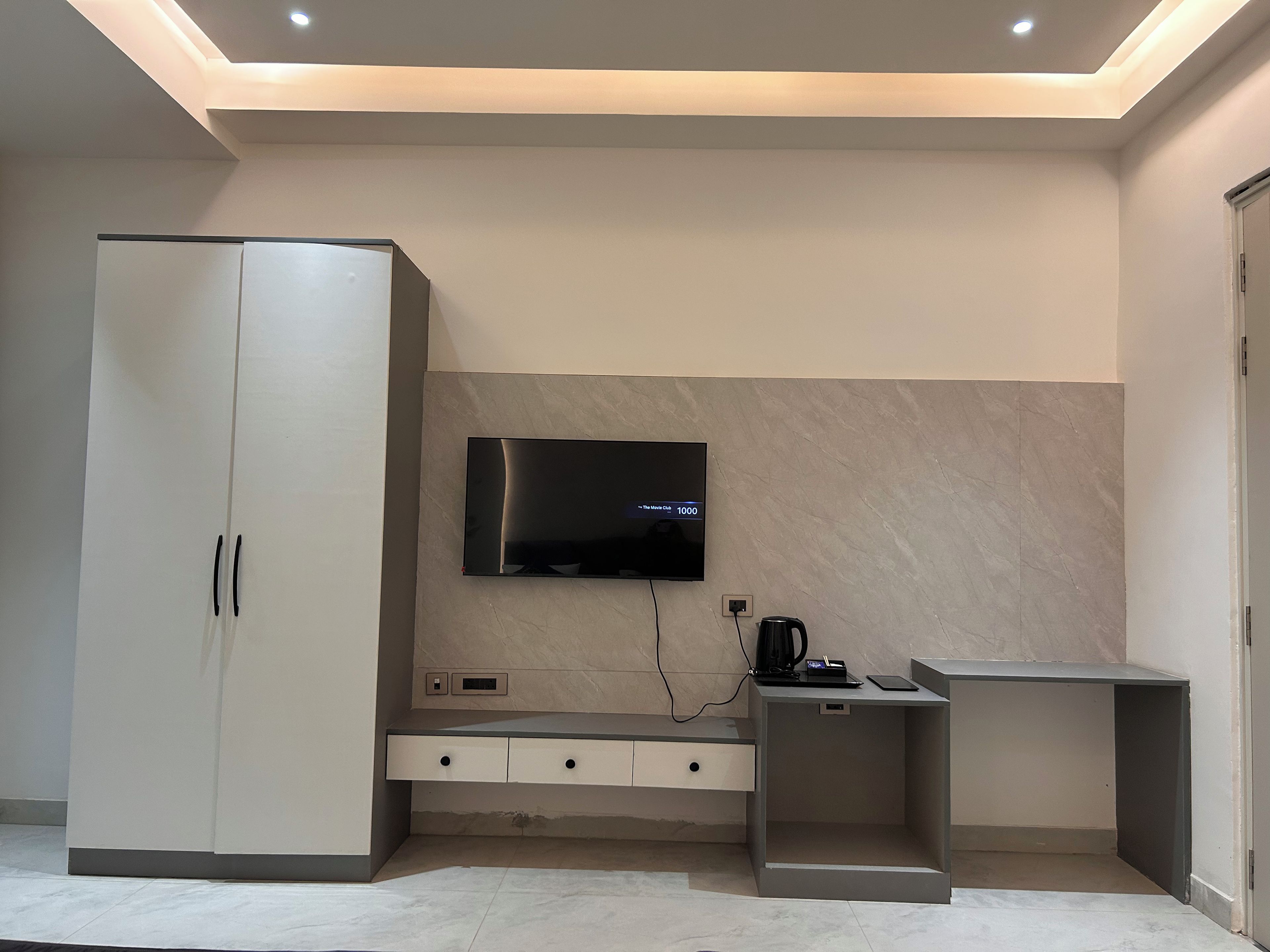 Quarto moderno com televisão na parede, guarda-roupa, secretária com cadeira e máquina de café.