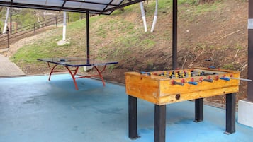 Mesa de futebol de mesa e mesa de pingue-pongue em área coberta, com vista para uma encosta.