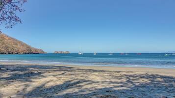 Uma praia com céu claro, mar calmo e barcos ao longe.