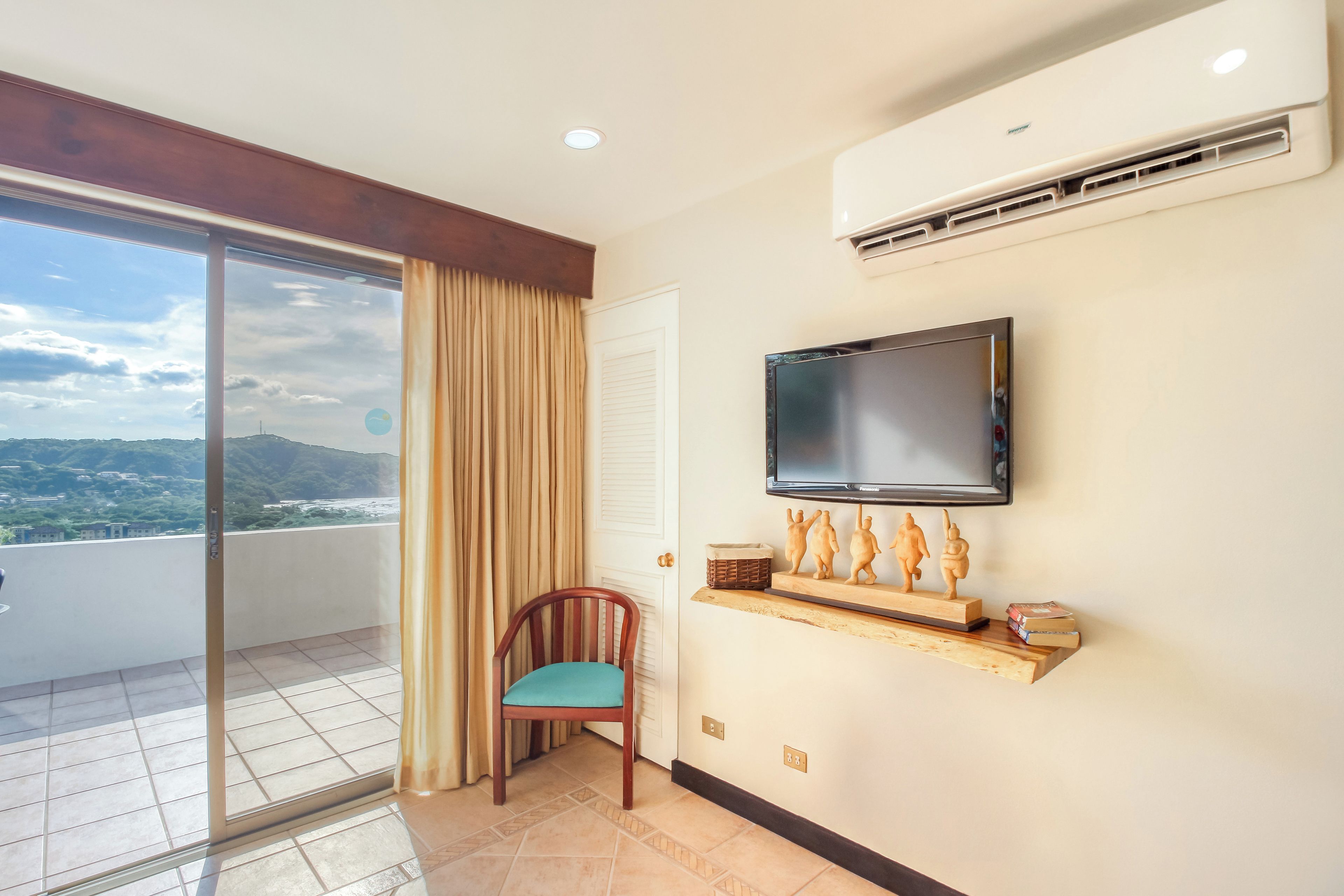 Quarto com televisão na parede, uma cadeira, prateleira de madeira com figuras e porta de vidro deslizante que dá para uma varanda com vista para a paisagem.