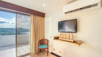 Quarto com televisão na parede, uma cadeira, prateleira de madeira com figuras e porta de vidro deslizante que dá para uma varanda com vista para a paisagem.