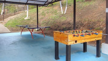 Espaço ao ar livre com mesa de futebol de mesa e mesa de pingue-pongue sob uma estrutura coberta.
