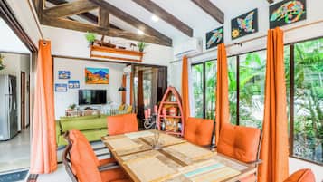 Sala de estar moderna com uma televisão grande, área de jantar com cadeiras laranja e vista para vegetação tropical pelas janelas.