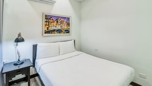 Quarto com cama, mesa de cabeceira, abajur, ar condicionado e uma pintura de paisagem urbana.