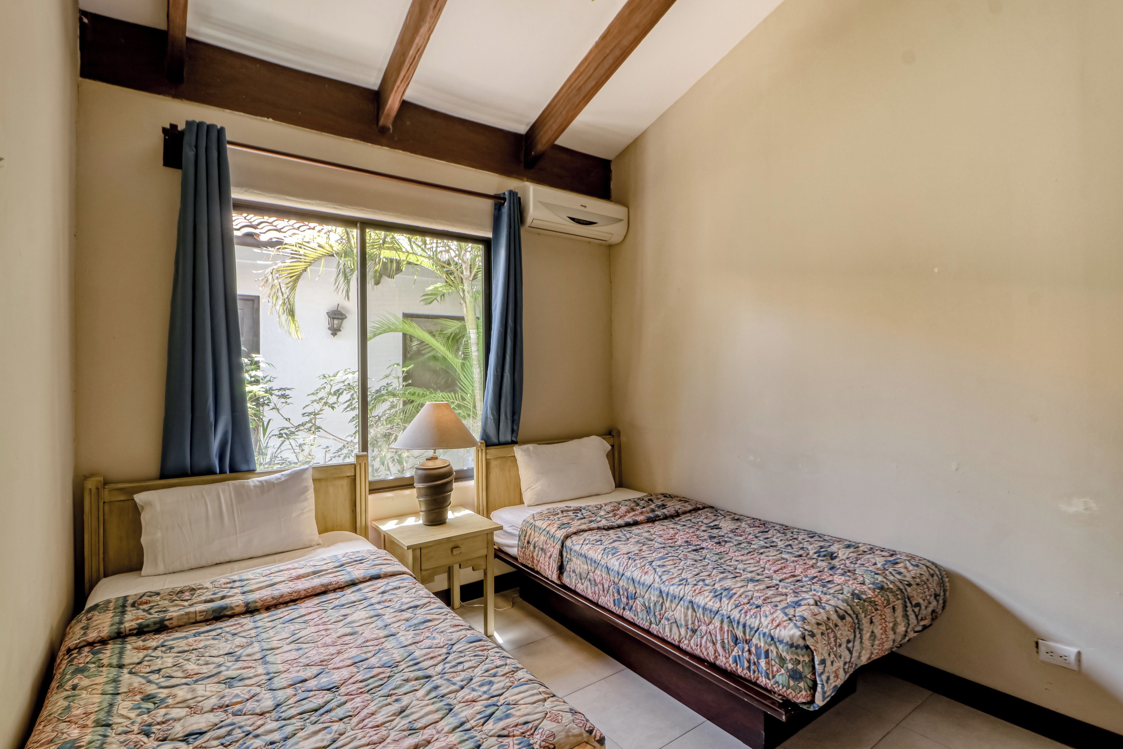 Quarto com duas camas, janela com cortinas, ar condicionado e abajur em uma mesinha de cabeceira.