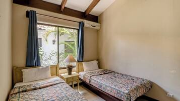 Quarto com duas camas, janela com cortinas, ar condicionado e abajur em uma mesinha de cabeceira.