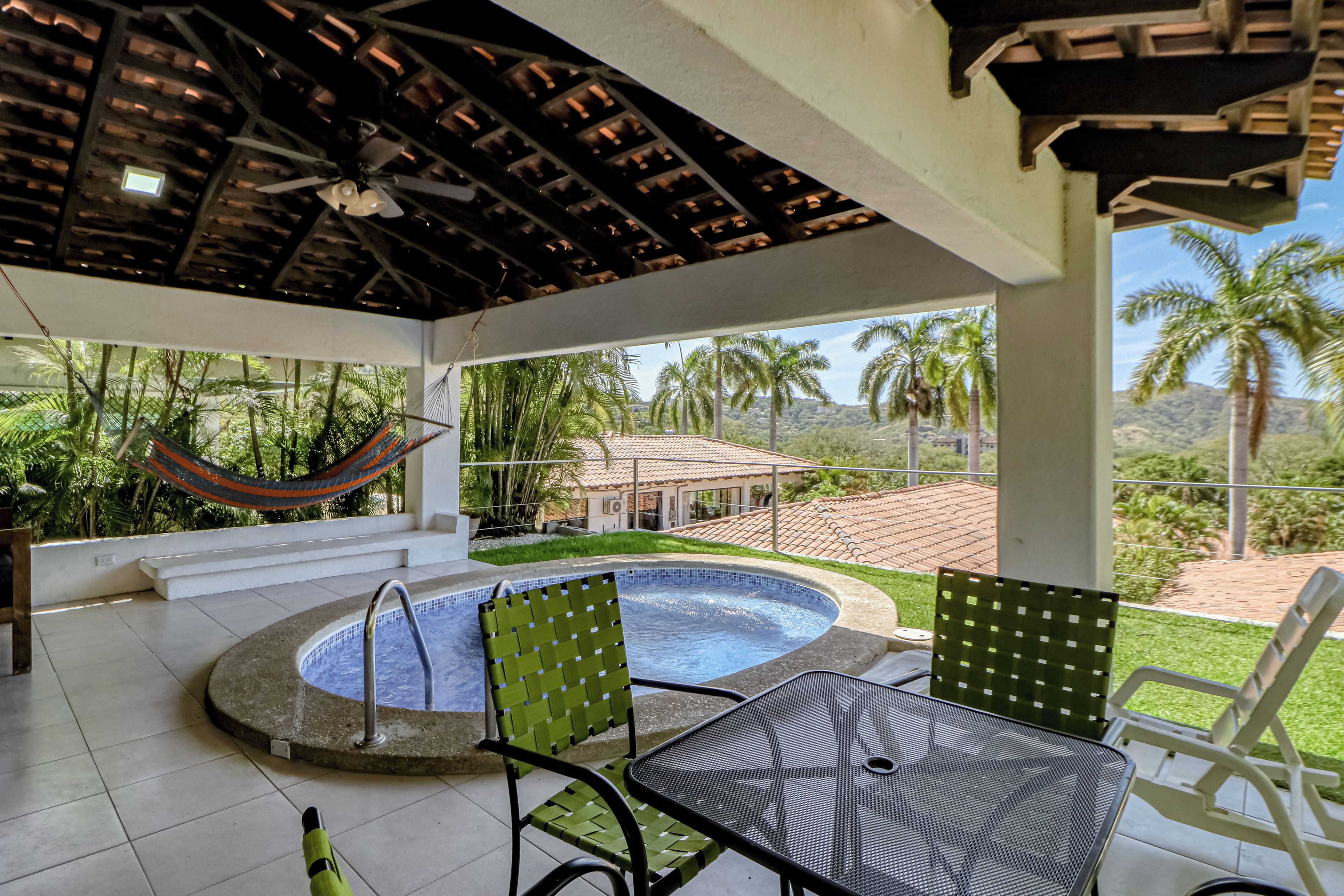 Área externa coberta com piscina, assentos e vista para palmeiras e uma casa.