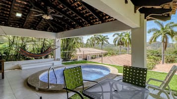 Área externa coberta com piscina, assentos e vista para palmeiras e uma casa.