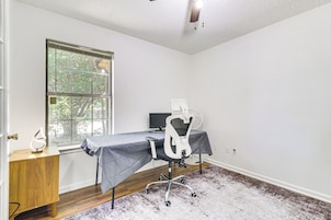 Quarto com escrivaninha de madeira, cadeira, computador e janela com vista para árvores.