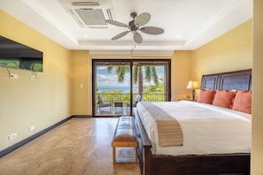 Quarto de hotel com cama, um banco, ventilador de teto, TV fixada na parede e vista para o exterior por uma porta de correr.