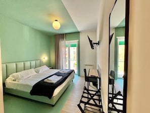 Quarto de hotel com cama, escrivaninha e cadeira. Há uma janela com cortinas, um radiador e uma luminária fixada na parede.