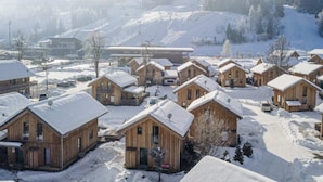 Um conjunto de cabanas de madeira com telhados cobertos de neve, situado em uma paisagem nevada com montanhas ao fundo.