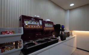 Uma estação de café com duas máquinas de expresso e uma cafeteira, um balcão de lanches com opções embaladas, e um micro-ondas em um balcão.
