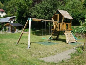 Estrutura de madeira para brincar com escorrega e balanços, em área gramada com uma casa e árvores ao fundo.