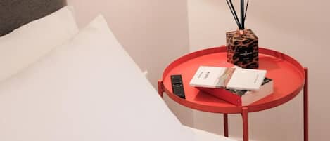 Cama bem arrumada com lençóis brancos, mesa de cabeceira vermelha com um livro e um controle remoto, e um vaso decorativo com gravetos pretos.