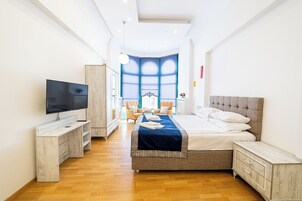 Quarto de hotel moderno com uma cama grande, televisão, guarda-roupa e um espaço de estar com uma cadeira e mesa.