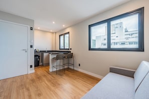 Apartamento moderno com cozinha compacta, área de jantar com banquetas, janela grande com vista para a cidade e sofá.