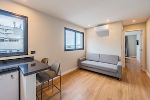 Cozinha moderna com geladeira embutida, espaço de refeições com um banco de bar, sofá cinza e vista para o horizonte urbano através de uma janela ampla.