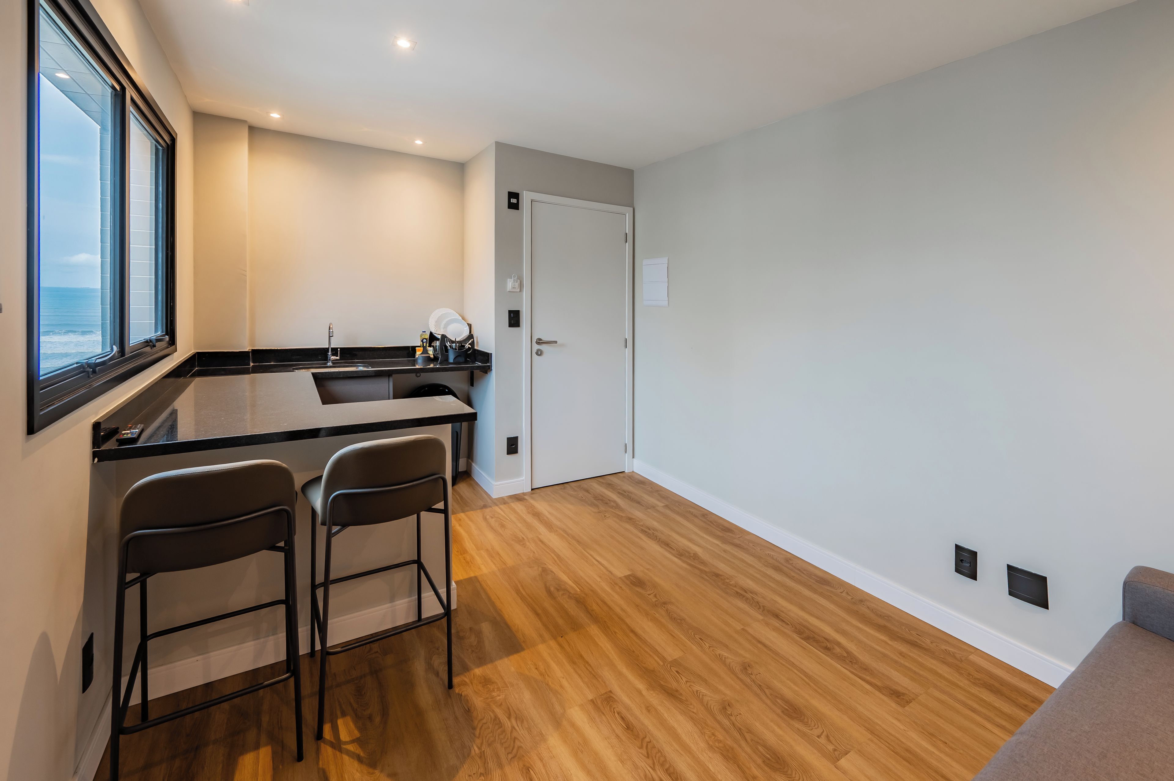 Cozinha moderna com bancada preta, dois banquetas e vista para o mar através de grandes janelas.