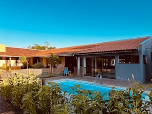 Área de piscina com céu azul límpido, alpendre coberto e um edifício com telhado em azulejos.