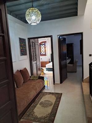Uma sala de estar com um sofá, um tapete estampado, um lustre pendurado e uma porta que dá para outro cômodo.