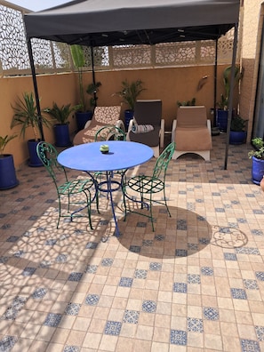 Uma área de varanda coberta com uma mesa redonda e cadeiras, plantas em vasos e um chão estampado.