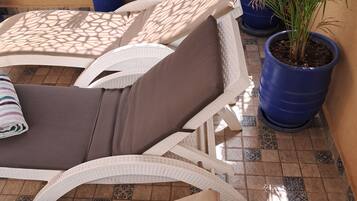 Uma área externa ensolarada com assentos de vime branco e almofadas, cercada por plantas em vasos.