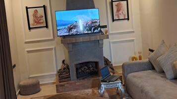 Uma sala de estar com lareira, televisão, sofá e um laptop sobre uma mesinha lateral.
